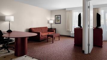 Quarto de hotel com carpete vermelho, sofá, mesa pequena, escrivaninha com cadeira, luminária, quadro na parede e televisão sobre um móvel.