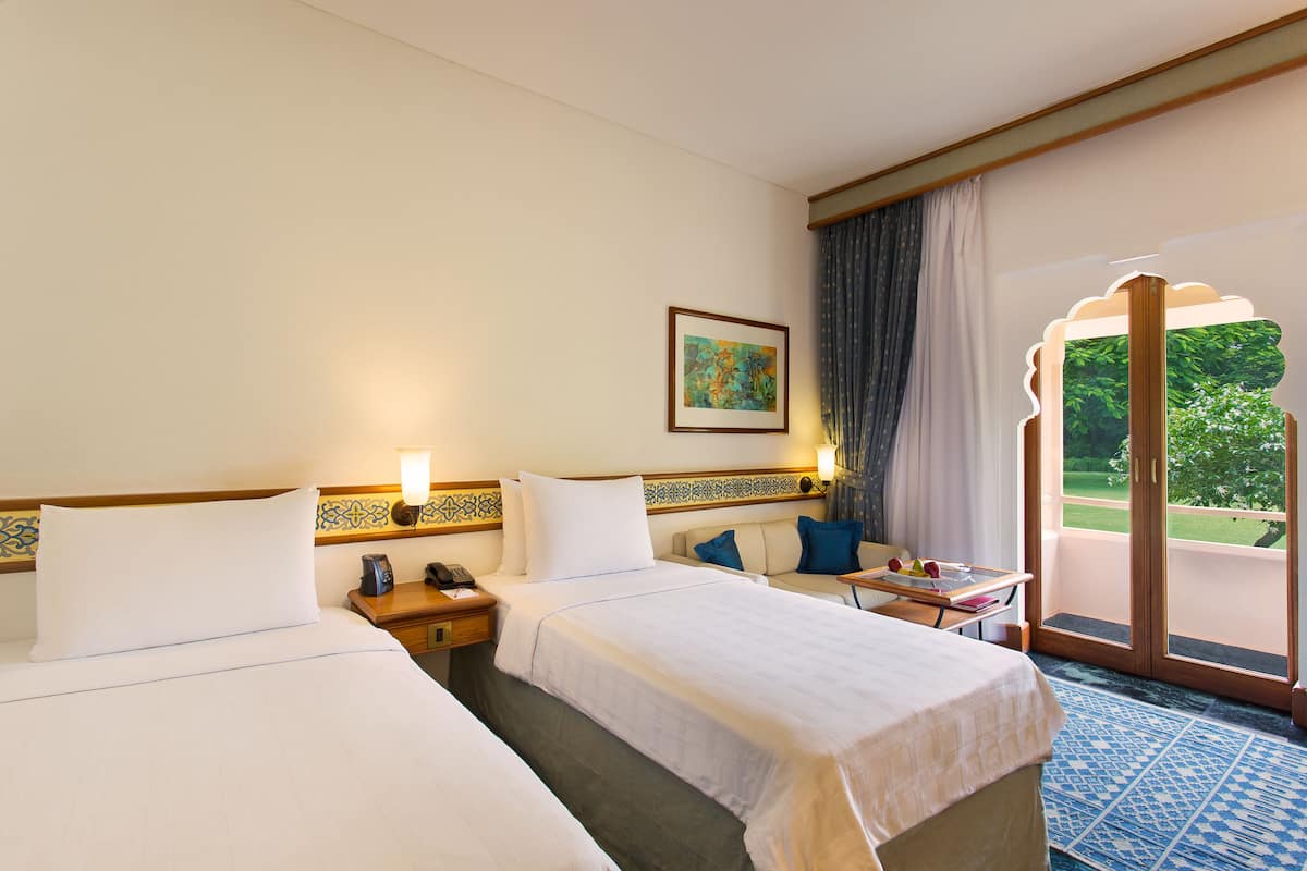 Quarto de hotel com duas camas, uma mesinha com bandeja, um quadro na parede e vista para o verde pela janela.