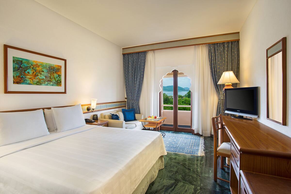 Quarto de hotel com uma cama, uma mesa com televisão, uma cadeira, uma pequena mesa com frutas e vista para o exterior por uma porta de vidro.