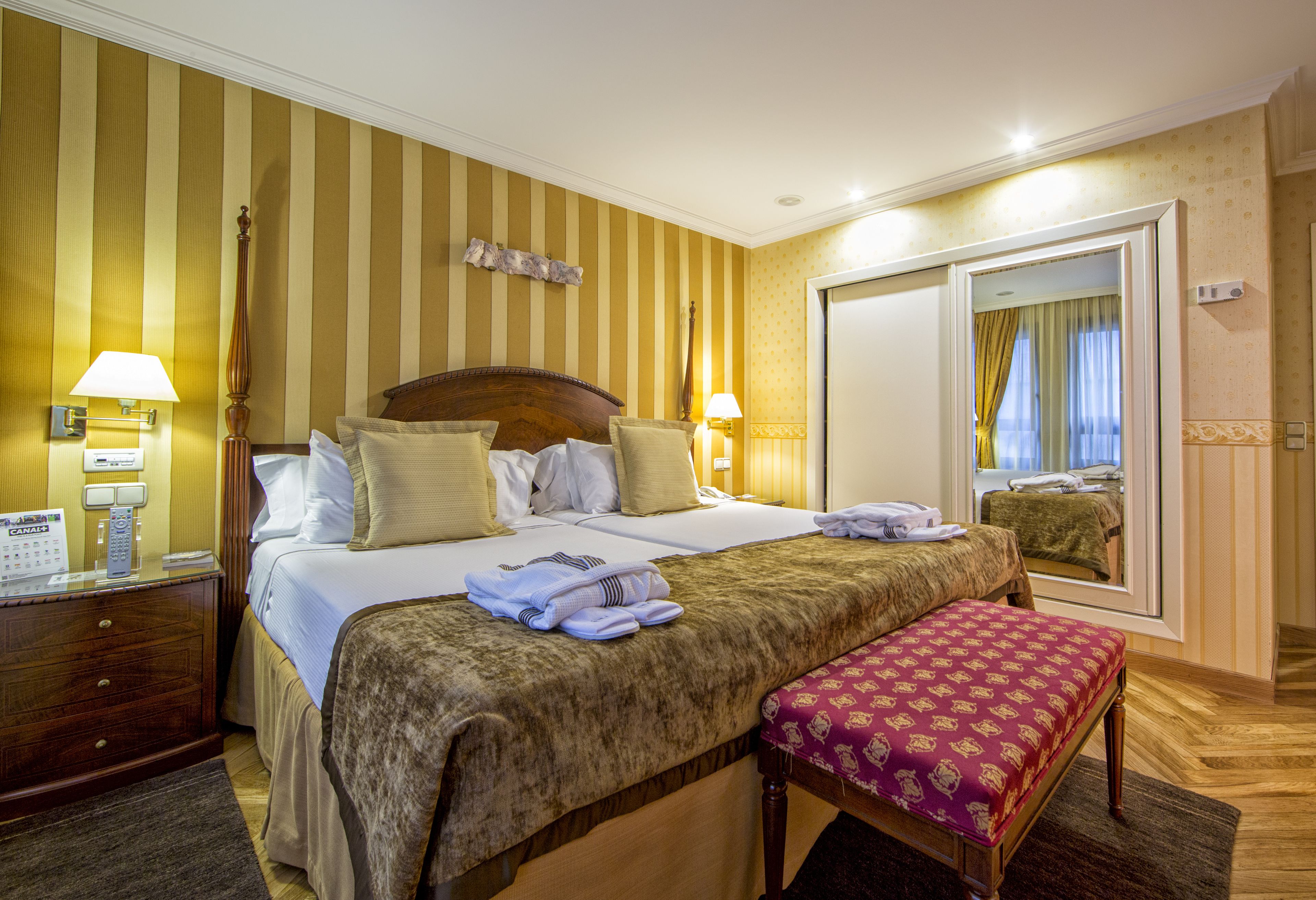 Quarto de hotel com uma cama grande, duas mesas de cabeceira com luminárias, um banco e um espelho.
