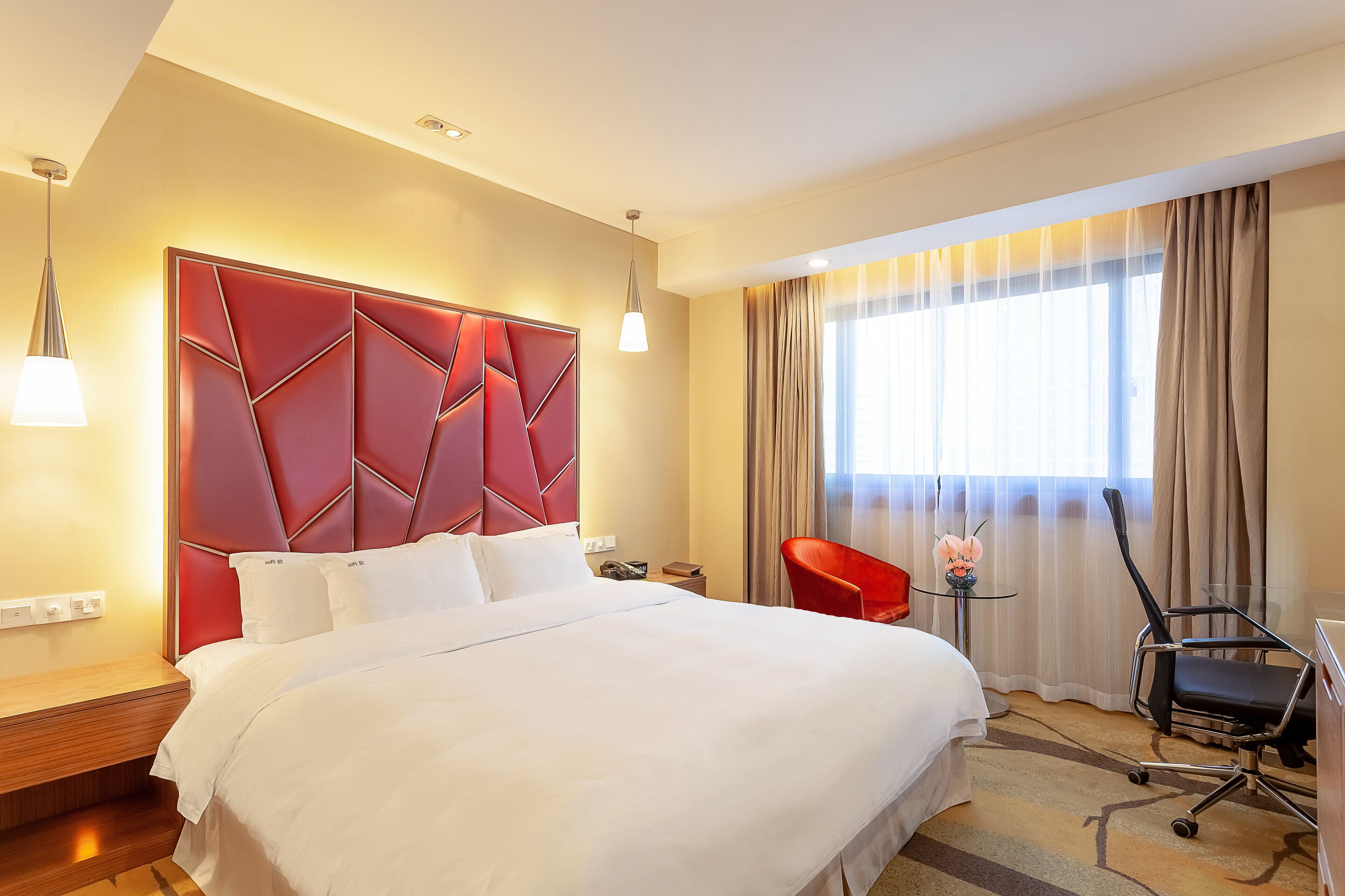 Quarto de hotel com uma cama grande, cabeceira vermelha, escrivaninha com cadeira e janela com cortinas.