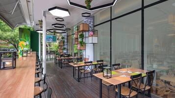 Interior moderno de café com mesas e cadeiras de madeira, luminárias de teto geométricas e plantas em vasos.