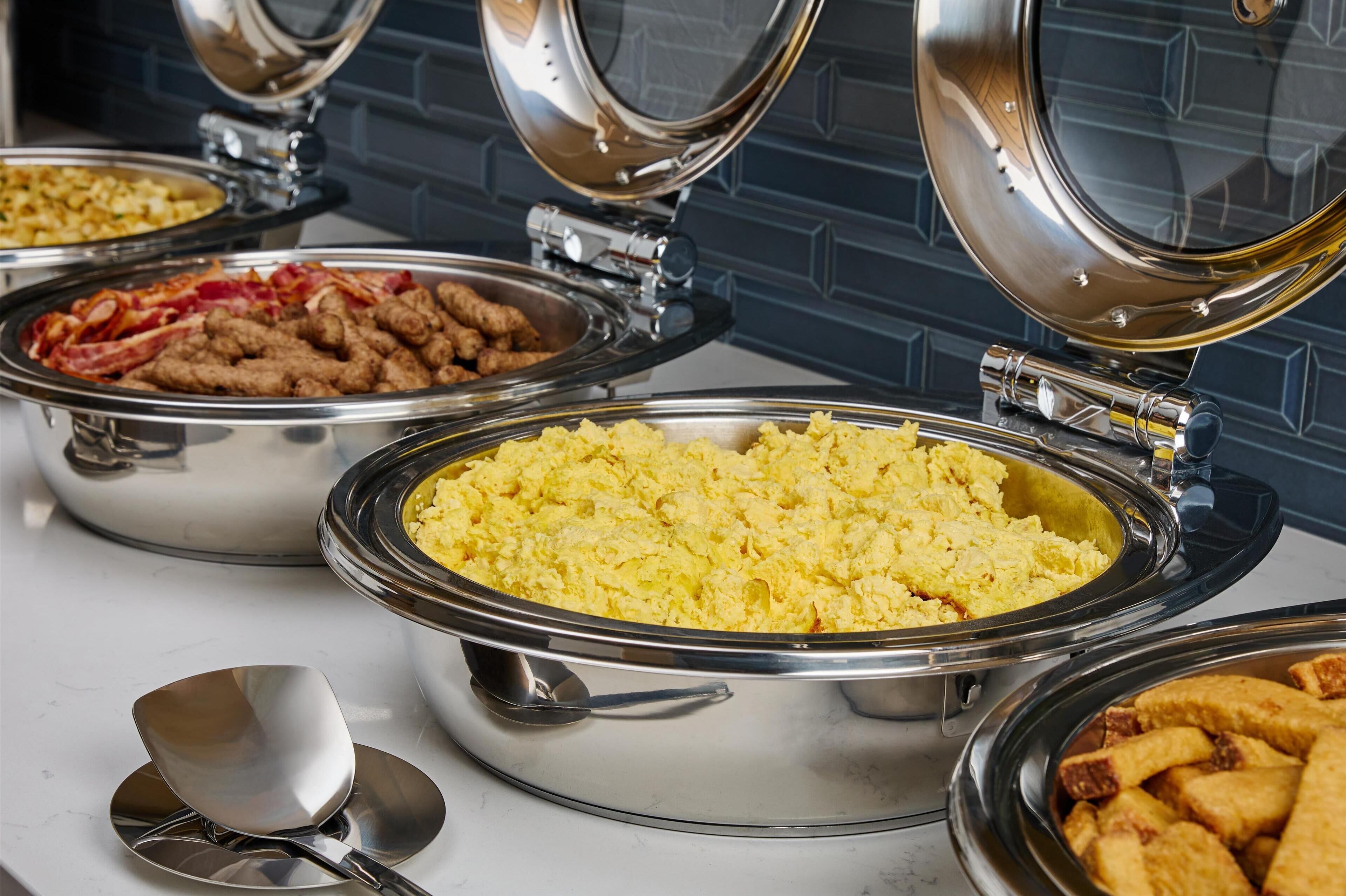 Buffet com diversos alimentos em recipientes de aço inoxidável, incluindo ovos mexidos, bacon e batatas fritas.