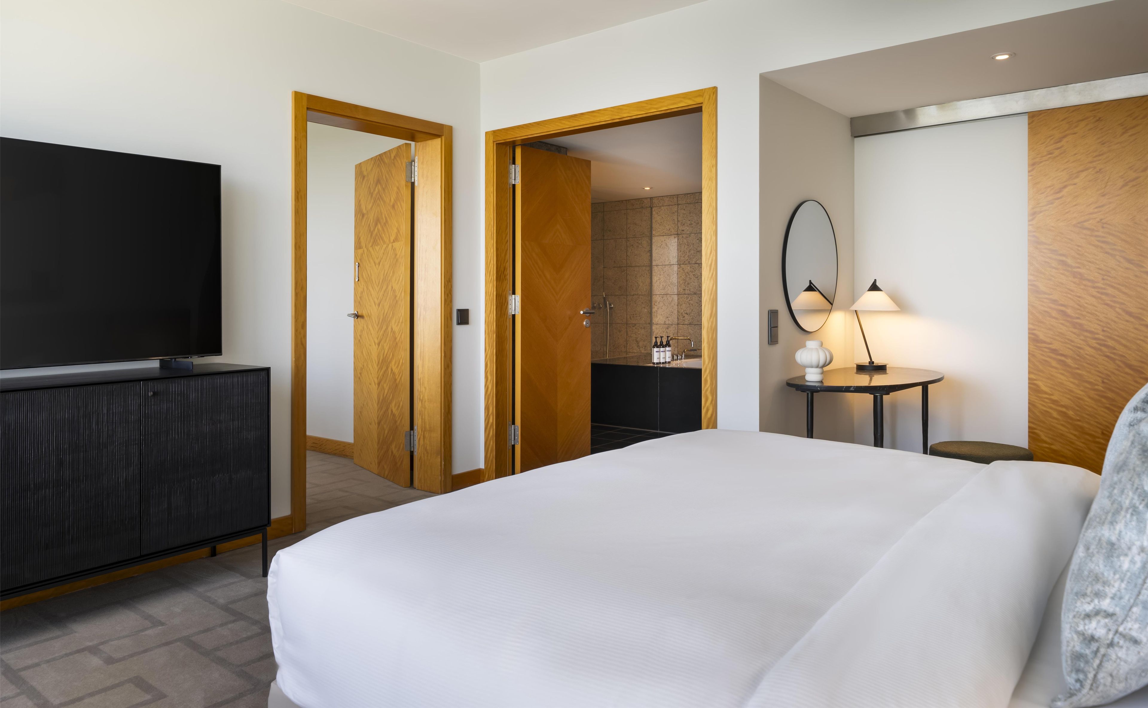 Quarto de hotel com cama, televisão, banheiro e uma pequena mesa com abajur.