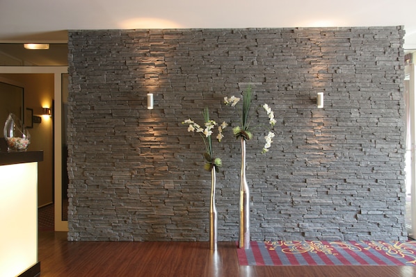 Uma área de recepção com parede de pedra, dois vasos altos com flores e um balcão de madeira.