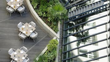 Terraço no telhado com várias mesas e cadeiras, cercado por vegetação exuberante e um edifício moderno com grandes janelas.