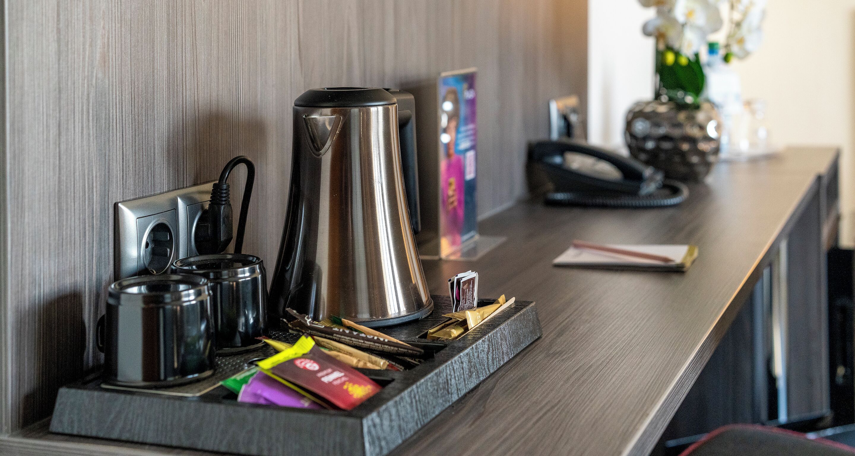 Bancada de recepção de hotel com cafeteira, telefone e uma pequena planta.