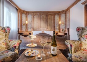 Quarto com cabeceira de madeira, poltronas florais, mesa de cabeceira com garrafa de vinho, copos e bandeja de pastelaria.