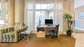 Quarto de hotel com escrivaninha, computador, cadeira, sofá e planta em vaso.
