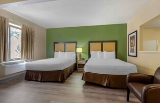 Quarto de hotel com duas camas, uma janela com cortinas, um aquecedor, mesa de cabeceira com abajur e uma poltrona de couro marrom.