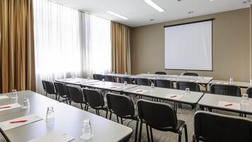 Sala de conferências com mesas, cadeiras, tela de projetor e uma grande janela com cortinas.