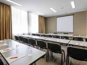 Sala de conferências com mesas, cadeiras, tela de projetor e uma grande janela com cortinas.