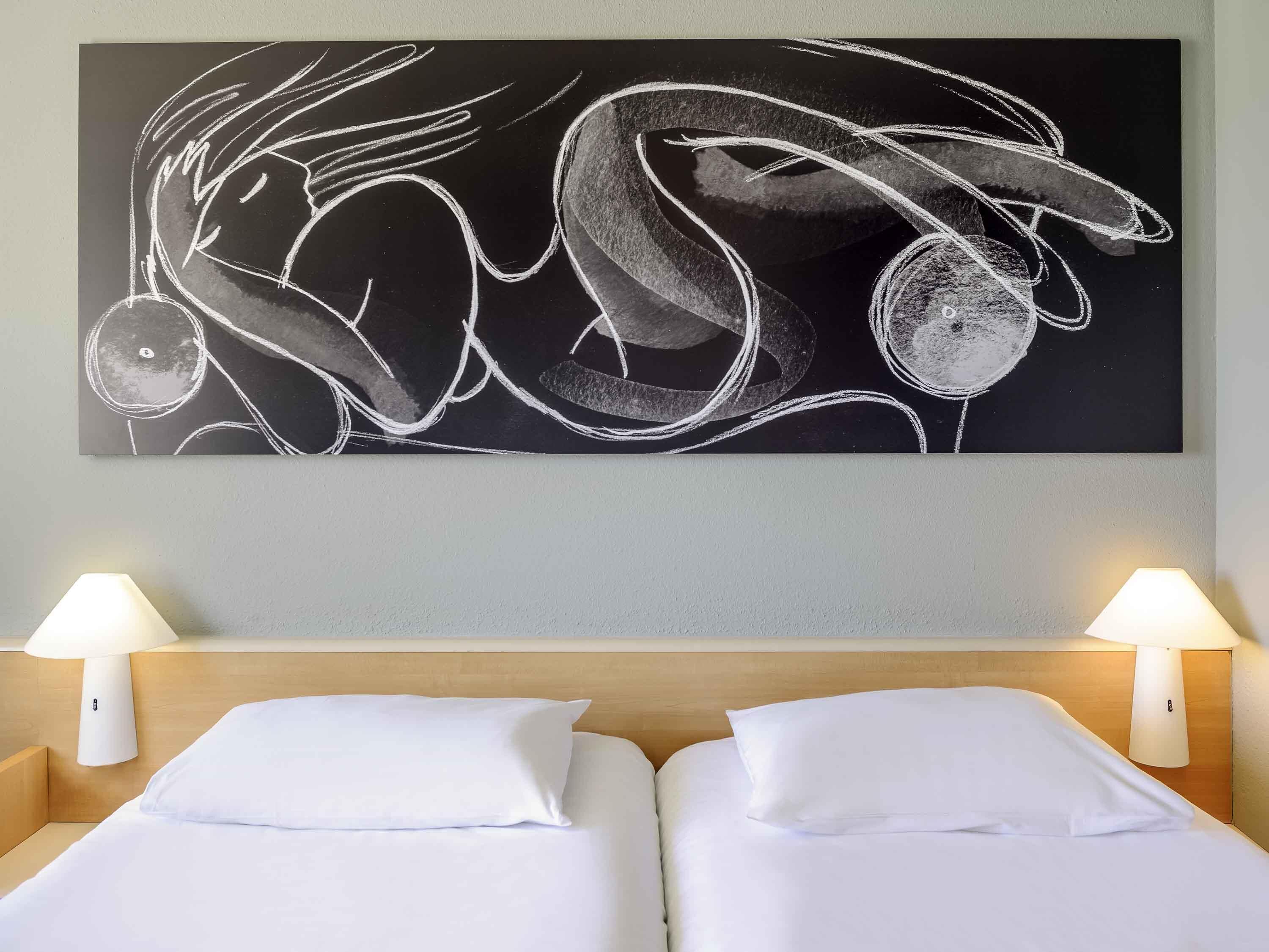 Quarto de hotel com uma grande obra de arte abstrata na parede, duas camas com roupa de cama branca e duas luminárias de cabeceira.