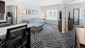 Quarto de hotel moderno com sofá cinzento, mesa de jantar com cadeiras, tapete com padrão preto e branco e uma televisão.