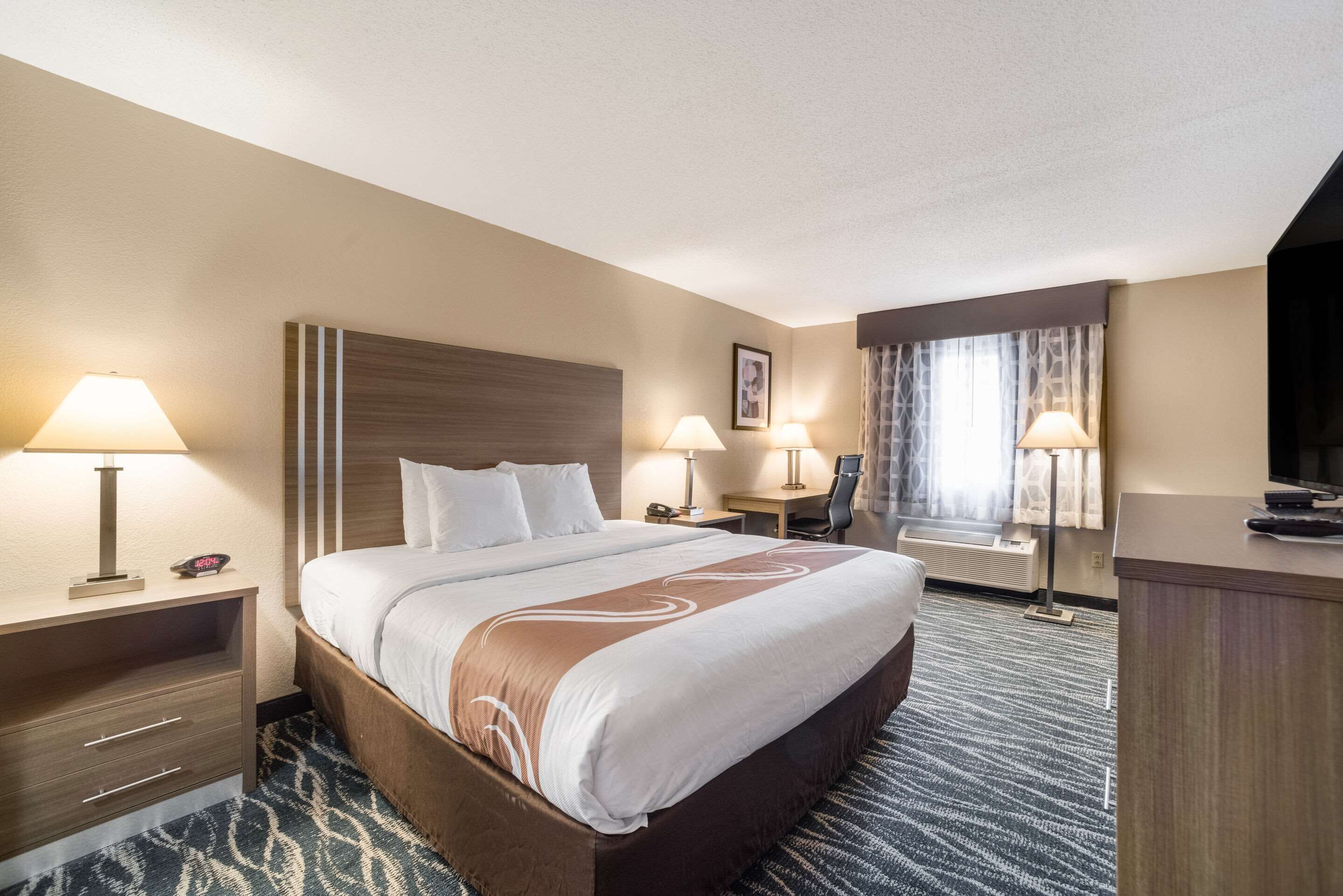 Quarto de hotel com uma cama grande, duas mesas de cabeceira com luminárias, uma escrivaninha, uma cadeira e uma televisão.