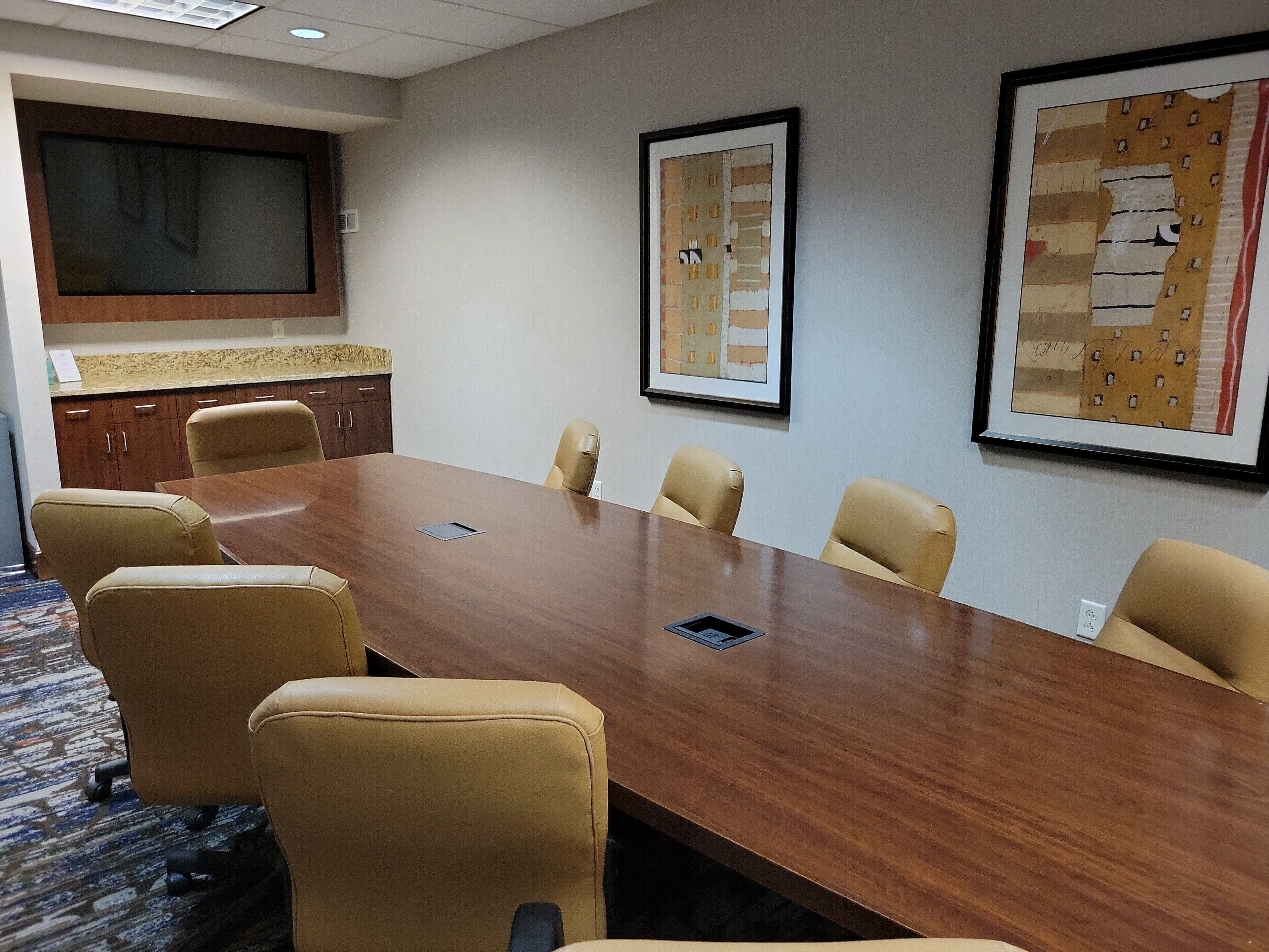 Sala de conferências com uma longa mesa de madeira, seis cadeiras bege, uma TV de tela plana na parede e duas obras de arte emolduradas nas paredes.