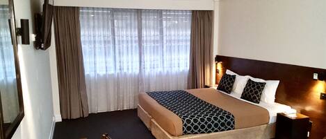 Quarto de hotel com uma cama grande, televisão, mesa de cabeceira e janela com cortinas.