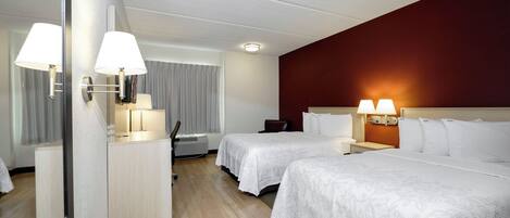 Quarto de hotel com duas camas, uma escrivaninha, uma cadeira, um espelho e uma janela com cortinas.