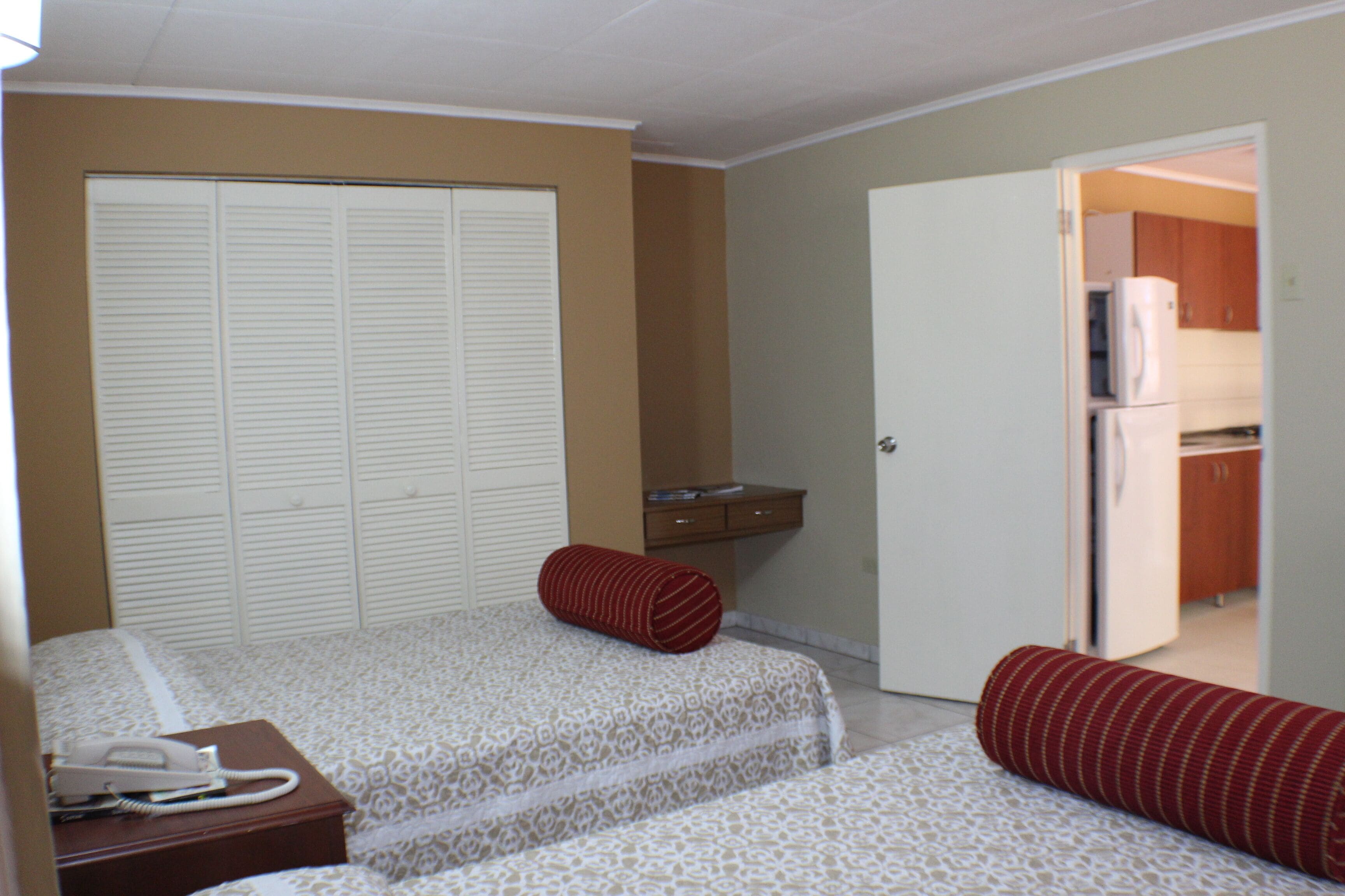 Quarto com duas camas, telefone ao lado da cama, guarda-roupa e uma kitchenette no quarto adjacente.