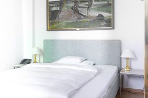 Cama bem arrumada com lençóis brancos, cabeceira cinza, abajur, telefone em uma mesa de cabeceira e um quadro na parede.