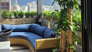 Varanda com sofá curvo, uma planta grande em vaso e vista para uma paisagem urbana com montanhas ao fundo.
