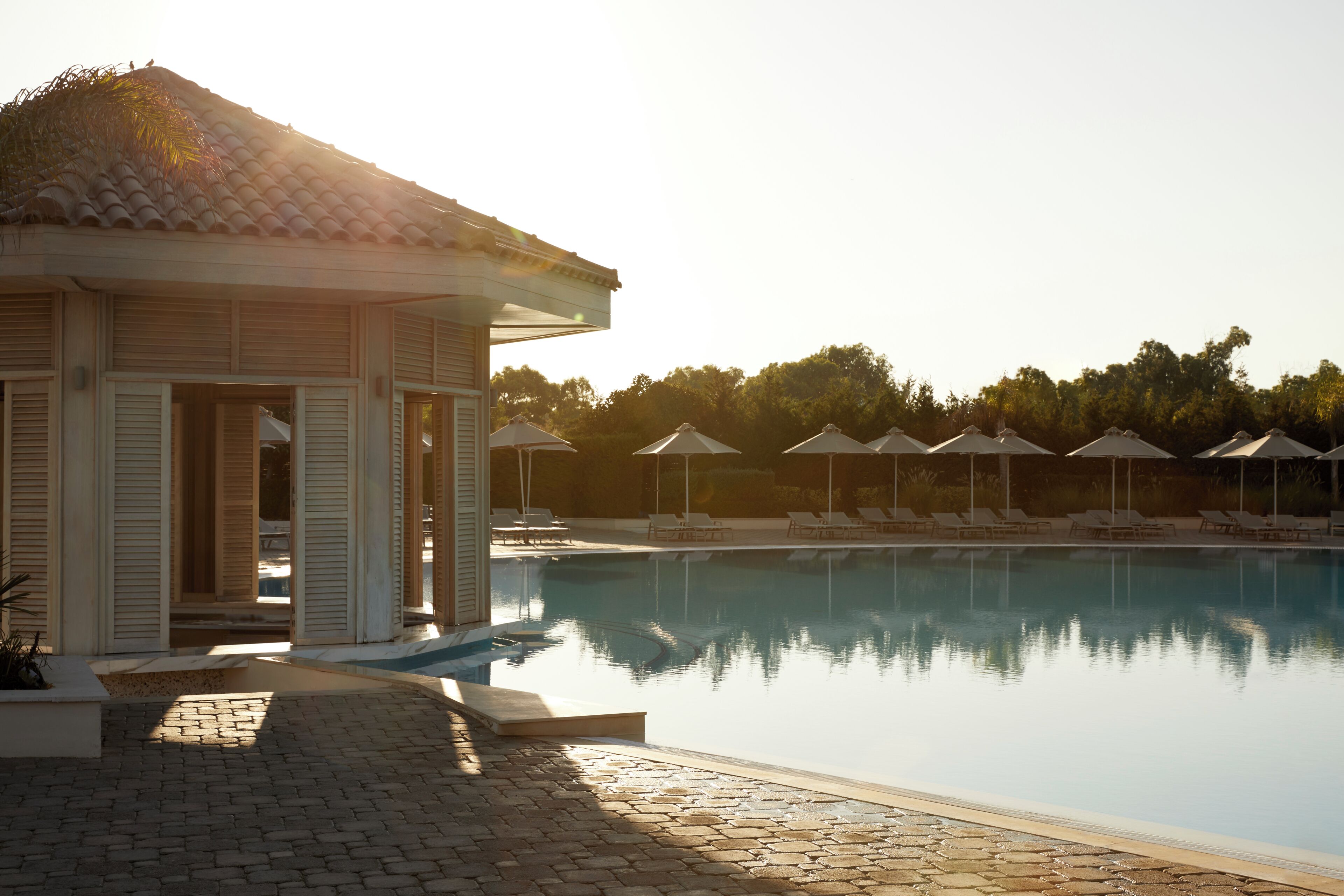 Piscina com espreguiçadeiras e guarda-sóis, um edifício com janelas abertas e céu claro.