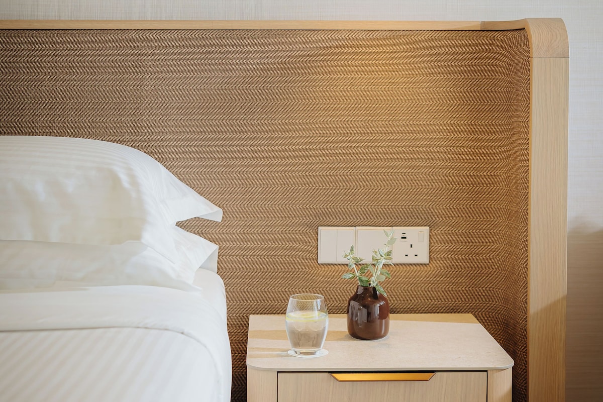 Uma cama bem arrumada com lençóis brancos, uma mesa de cabeceira com um vaso e um copo, e um lustre pendurado.