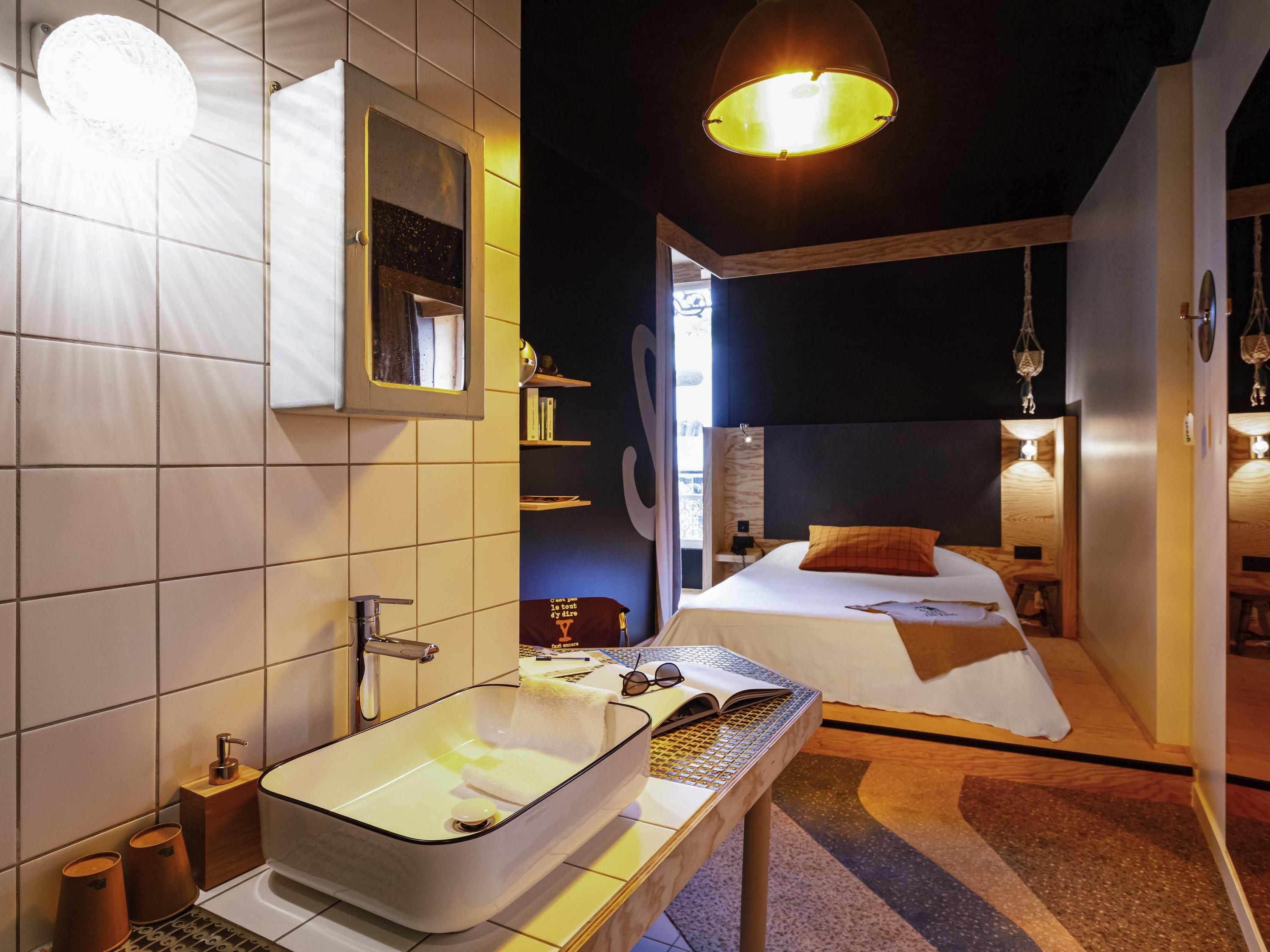 Quarto de hotel moderno com uma grande toucador de banheiro, uma cama com cabeceira de madeira e uma pequena mesa com um livro e copos.