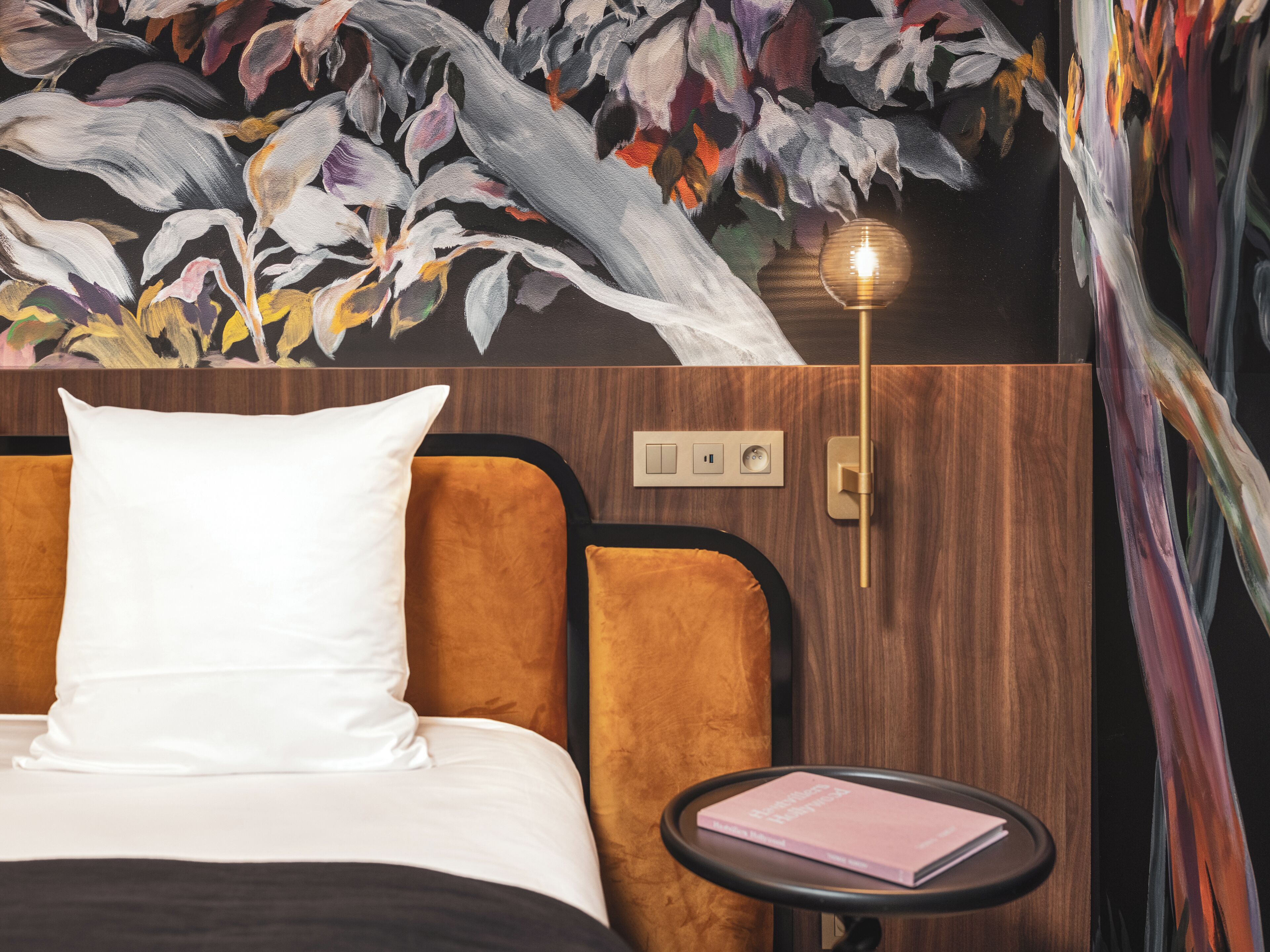 Quarto de hotel com uma cama, uma mesa de cabeceira com um livro, uma luminária na parede e uma tomada.