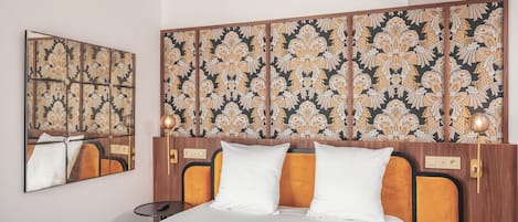 Quarto de hotel com uma cama grande, cabeceira com padrão decorativo, mesa de cabeceira com abajur e espelho na parede.