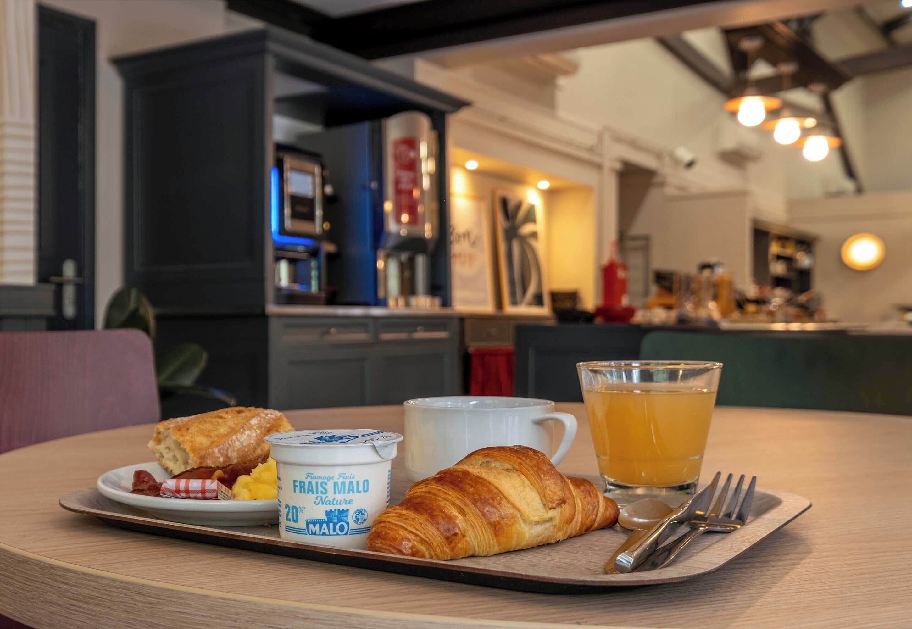 Bandeja de café da manhã com croissant, iogurte e suco de laranja sobre uma mesa de madeira em cozinha moderna.