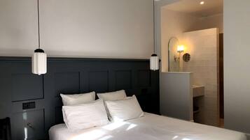 Quarto de hotel moderno com uma cama grande, duas luminárias pendentes, um banquinho pequeno e um banheiro visível por uma porta aberta.