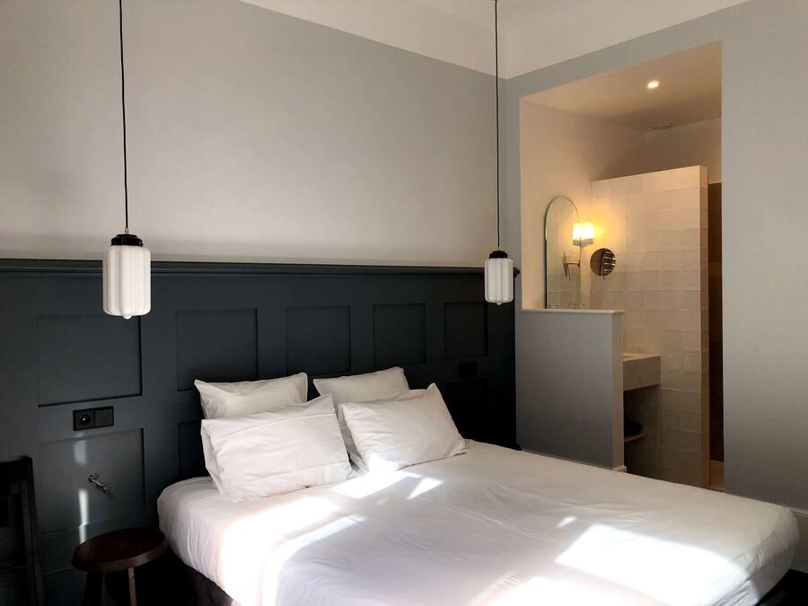 Quarto de hotel moderno com uma cama grande, duas luminárias pendentes, um banquinho pequeno e um banheiro visível por uma porta aberta.