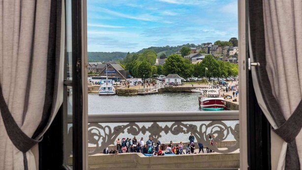 Vista pela janela com cortinas, mostrando um porto com barcos e uma multidão de pessoas.