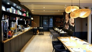 Um restaurante com um balcão comprido, várias mesas e cadeiras. Estantes com diversos objetos à esquerda.