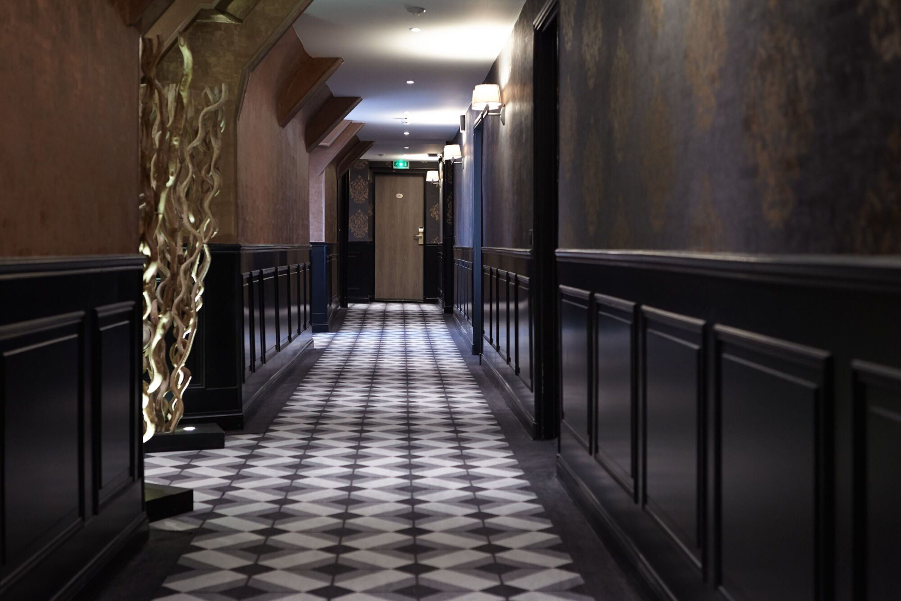 Um corredor com piso xadrez, paredes escuras e um elemento dourado decorativo à esquerda.