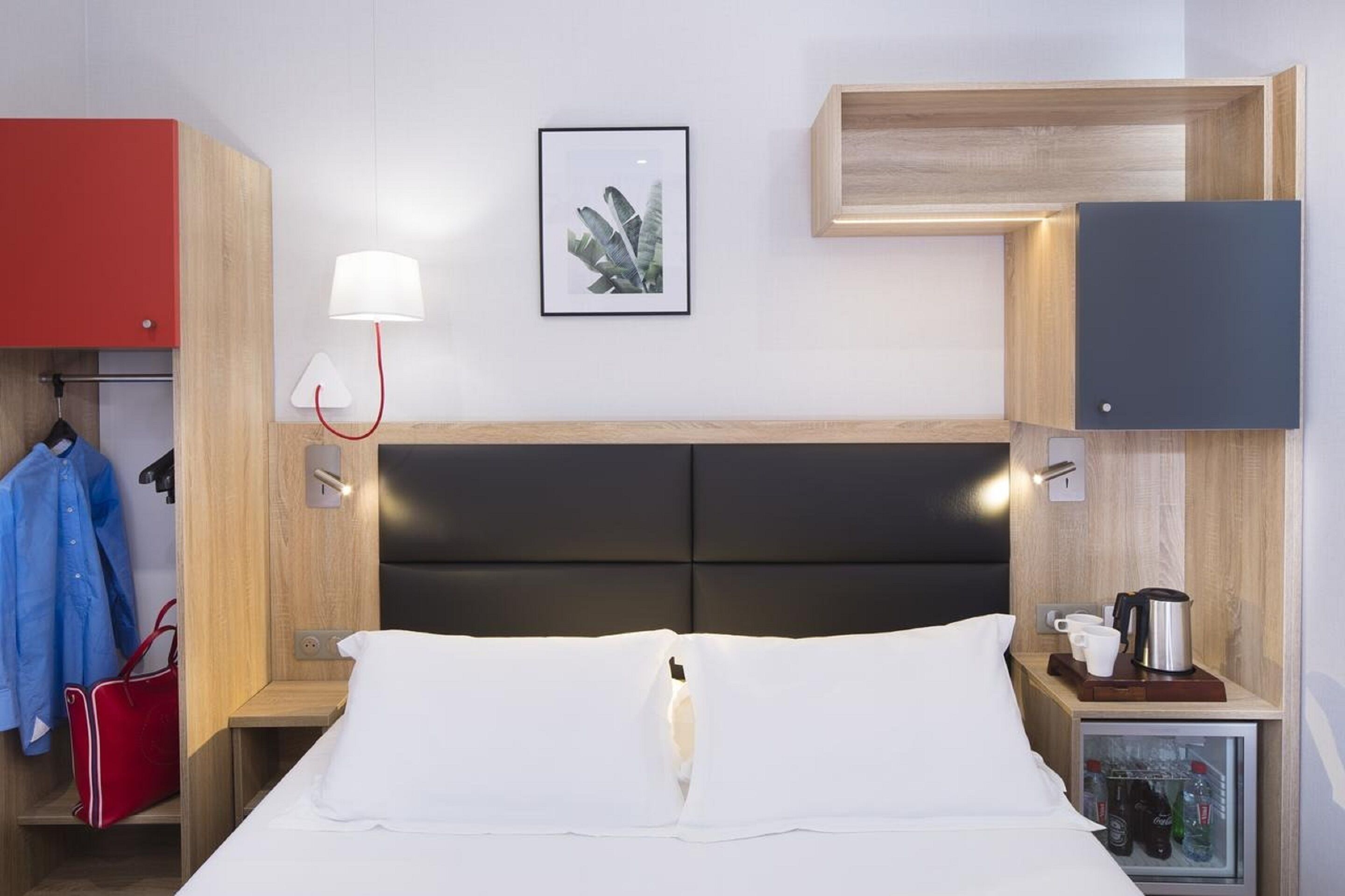 Quarto de hotel com cama, guarda-roupa, mesa de cabeceira com abajur, quadro emoldurado na parede e um pequeno armário.