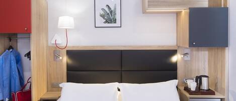 Quarto de hotel com cama, guarda-roupa, mesa de cabeceira com abajur, quadro emoldurado na parede e um pequeno armário.