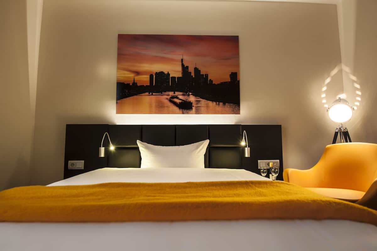 Quarto de hotel com cama, pintura de paisagem urbana, mesa de cabeceira, abajur e cadeira.