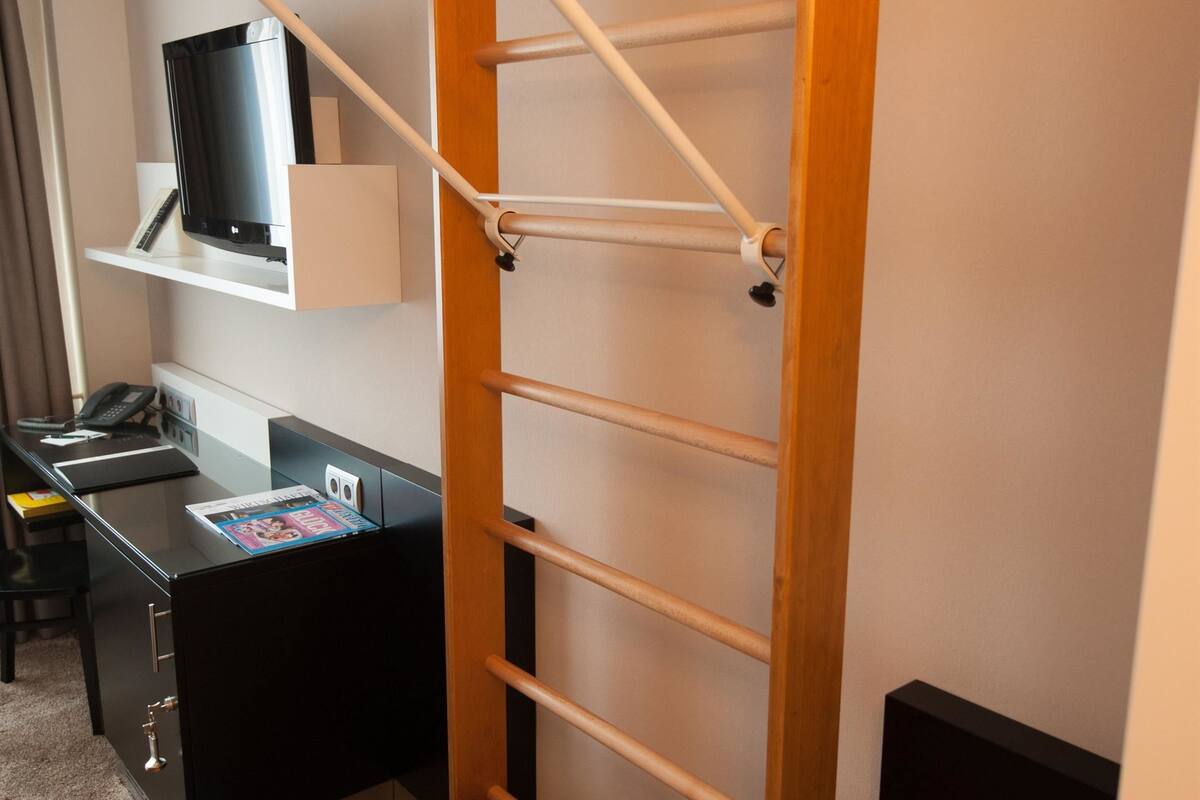 Quarto de hotel compacto com escrivaninha, televisão, estante e um banco de madeira.