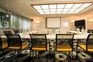 Uma sala de conferências com uma mesa comprida, cadeiras, um projetor e um amplo janelão com persianas.