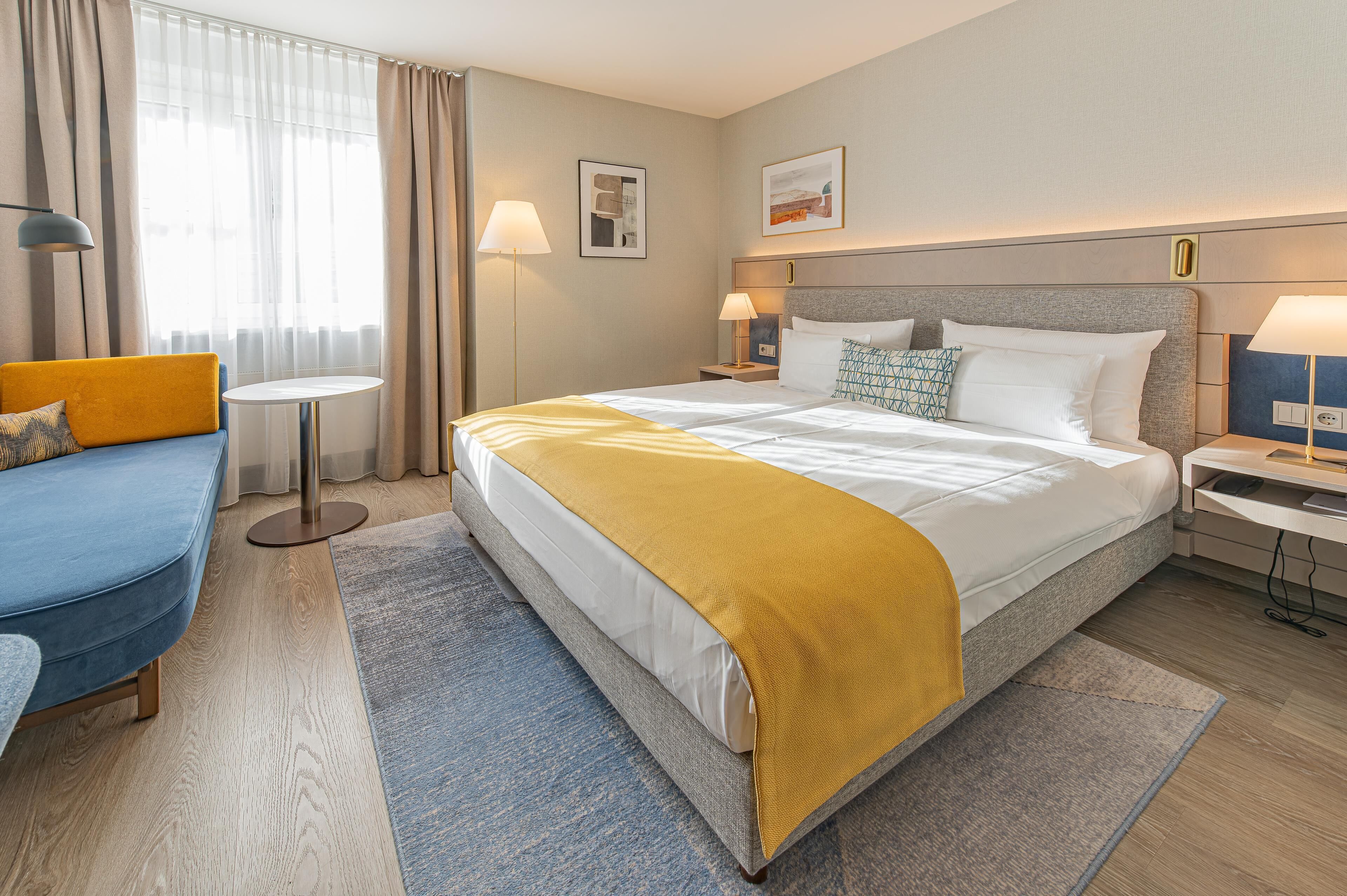 Quarto de hotel com cama, sofá, mesinha e abajur.