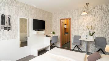 Quarto de hotel com cama, escrivaninha, duas cadeiras, televisão e porta para o banheiro.