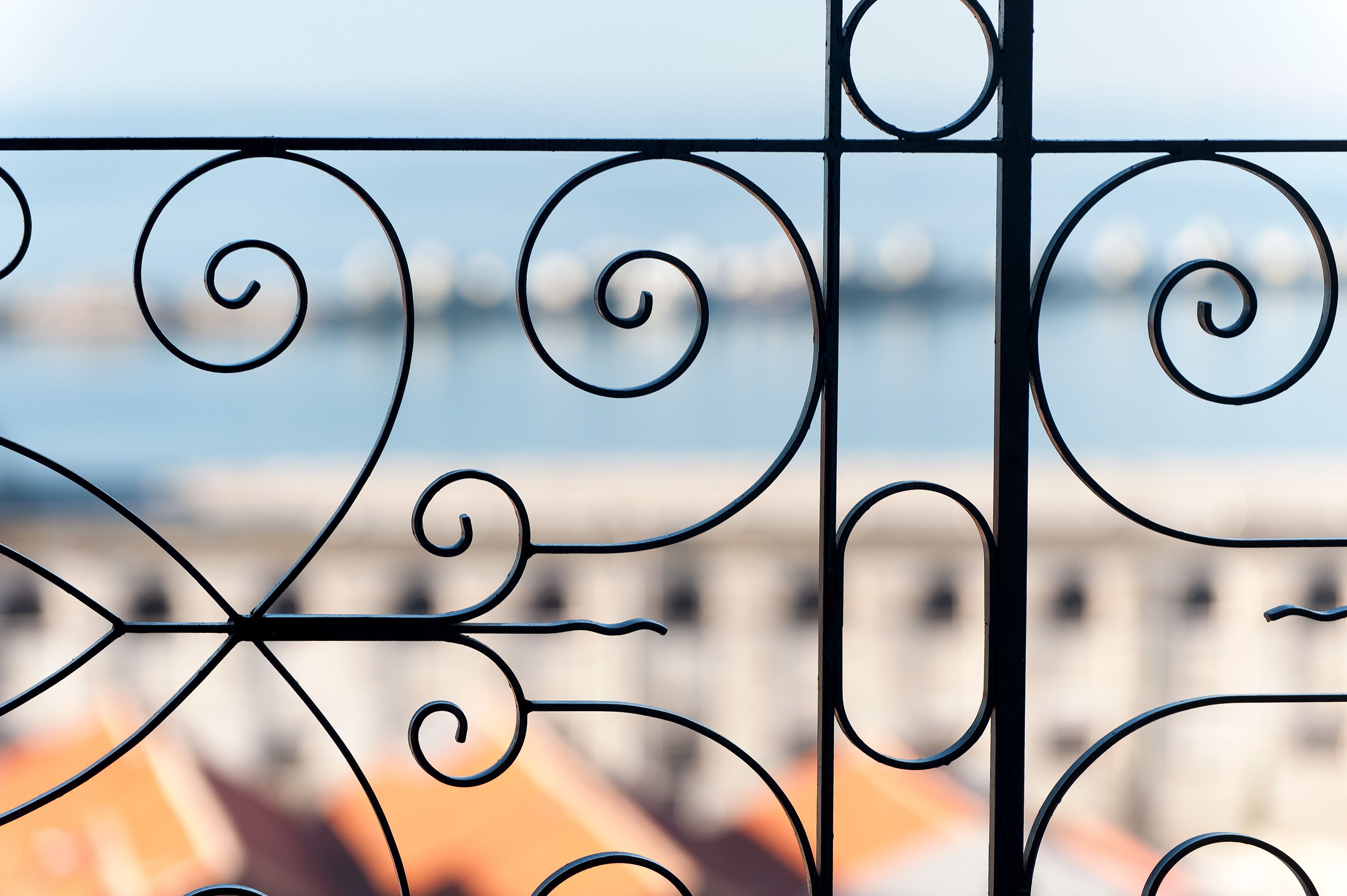 Close-up de um guarda-corpo metálico com desenhos elaborados, com vista para uma paisagem urbana com edifícios e um corpo de água ao fundo.