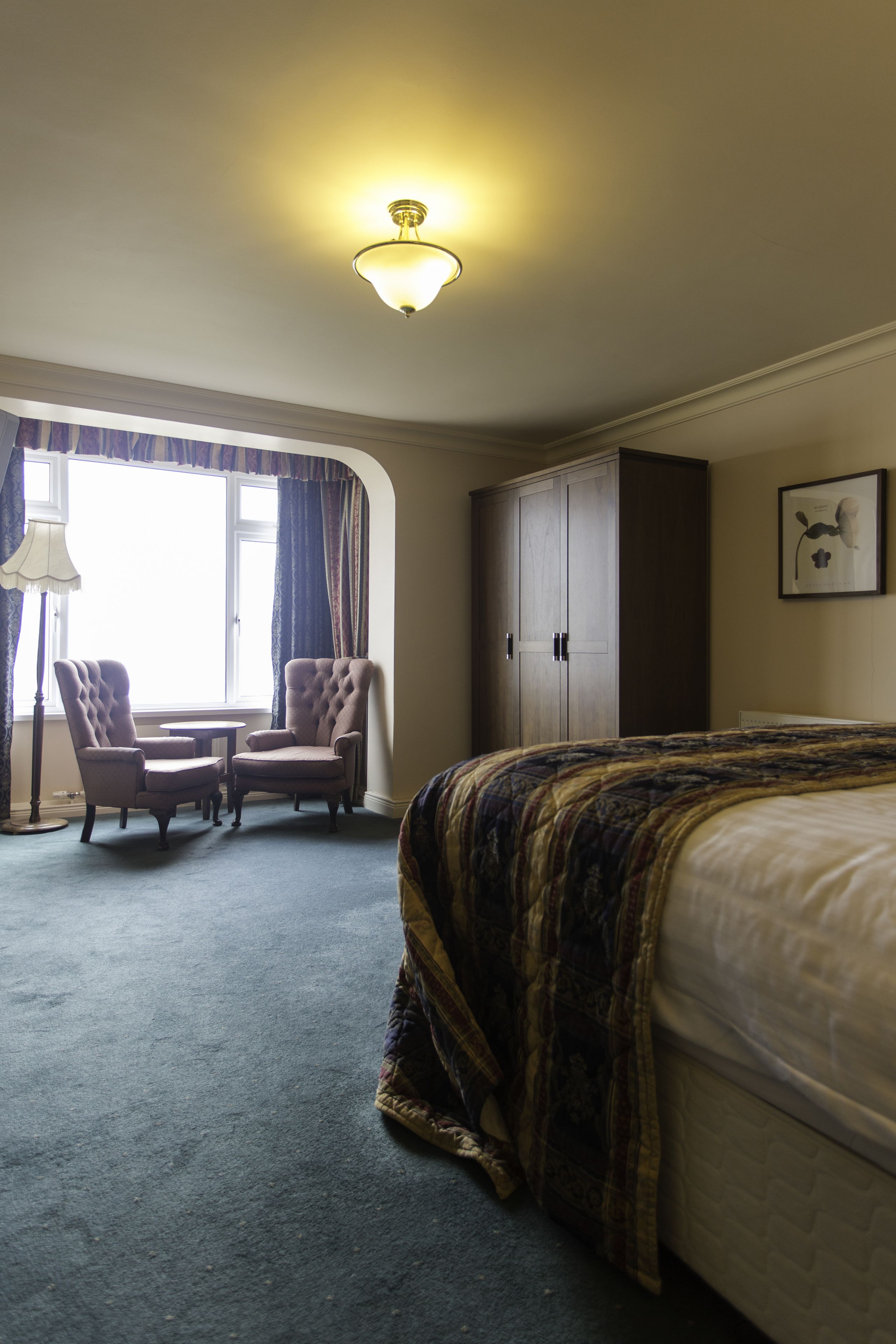 Quarto de hotel com uma cama grande, uma escrivaninha, duas cadeiras, um guarda-roupa e uma janela com cortinas.