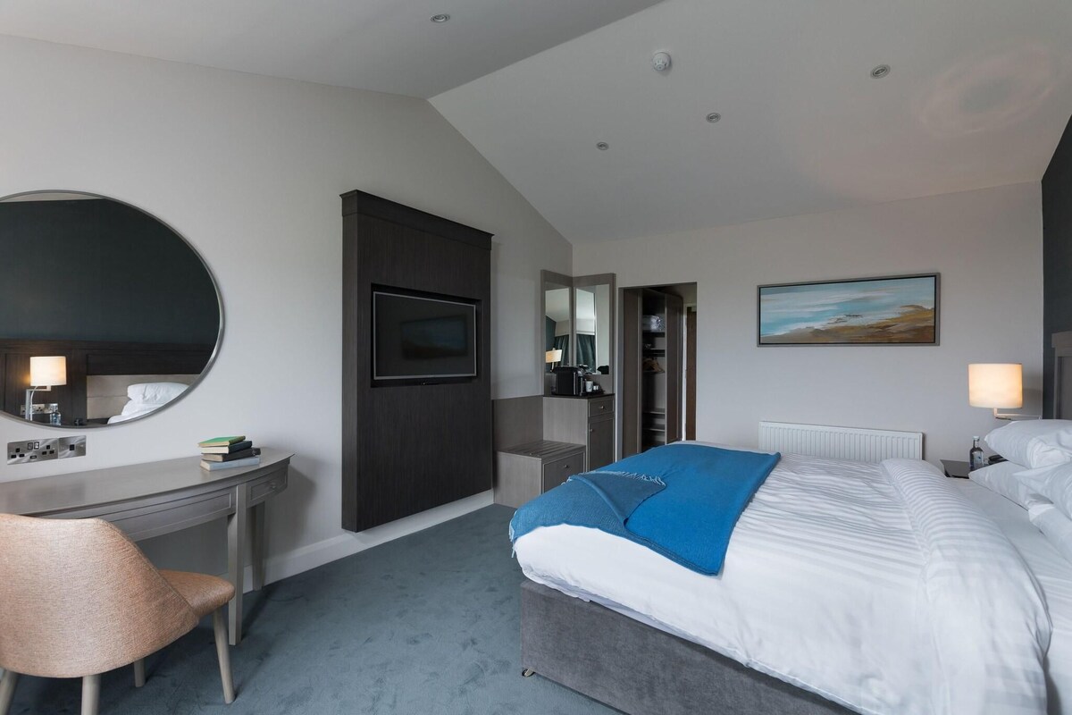 Quarto de hotel moderno com uma cama grande, uma escrivaninha com cadeira, televisão, um espelho amplo e um quadro na parede.