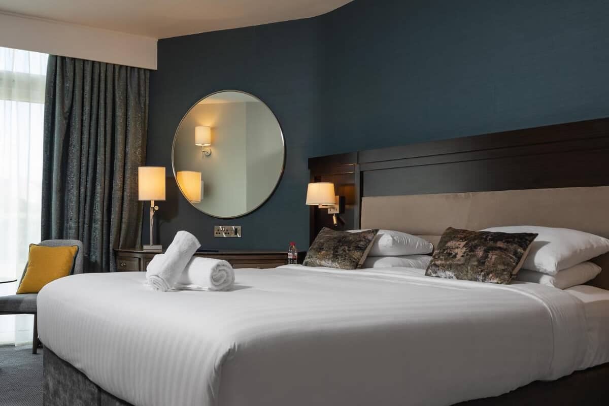 Quarto de hotel com uma cama grande, duas luminárias de cabeceira, um espelho redondo e vista para o exterior pela janela.