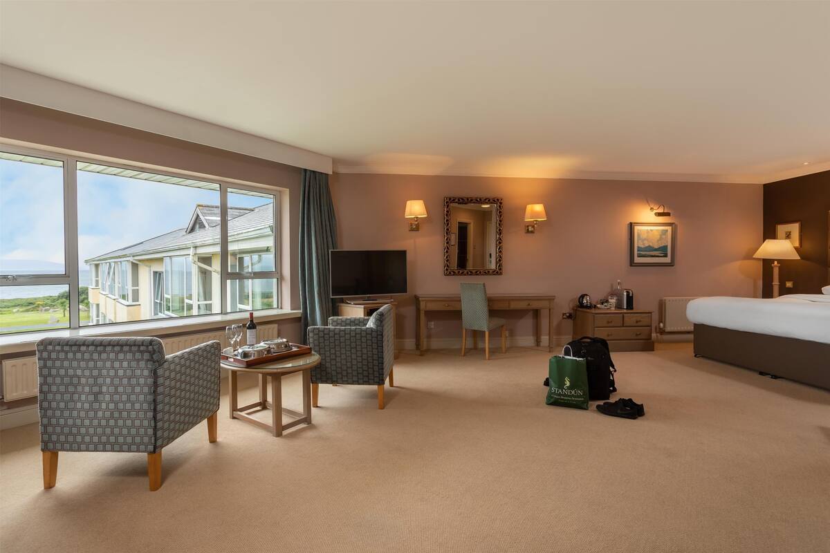 Quarto de hotel com uma grande janela, uma cama, uma mesa, uma cadeira, uma televisão e um abajur.