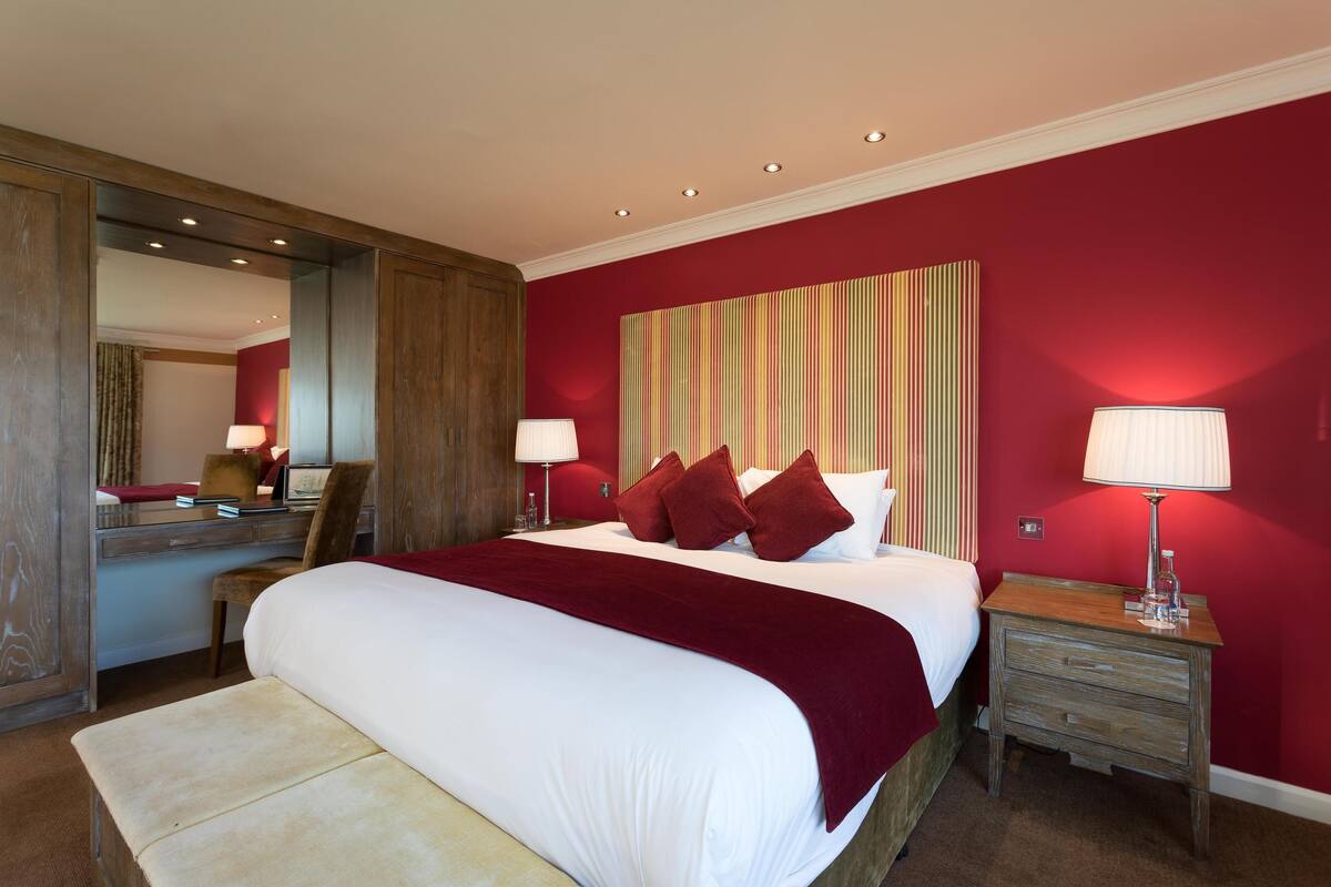 Quarto de hotel com uma cama grande, uma mesa de cabeceira de madeira com abajur, um espelho e cabeceira listrada.