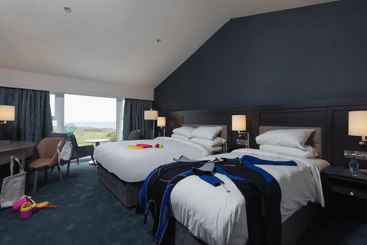 Quarto de hotel com uma cama grande, duas mesas de cabeceira, uma escrivaninha e uma cadeira. Janelas oferecem vista para o exterior.
